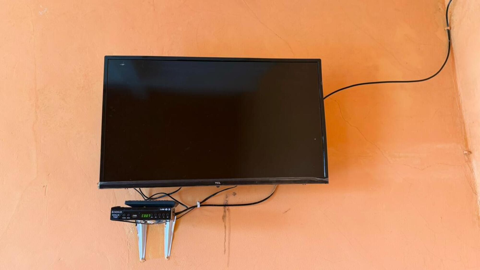 Uma TV de tela plana fixada na parede, com um decodificador e uma régua de tomadas logo abaixo.