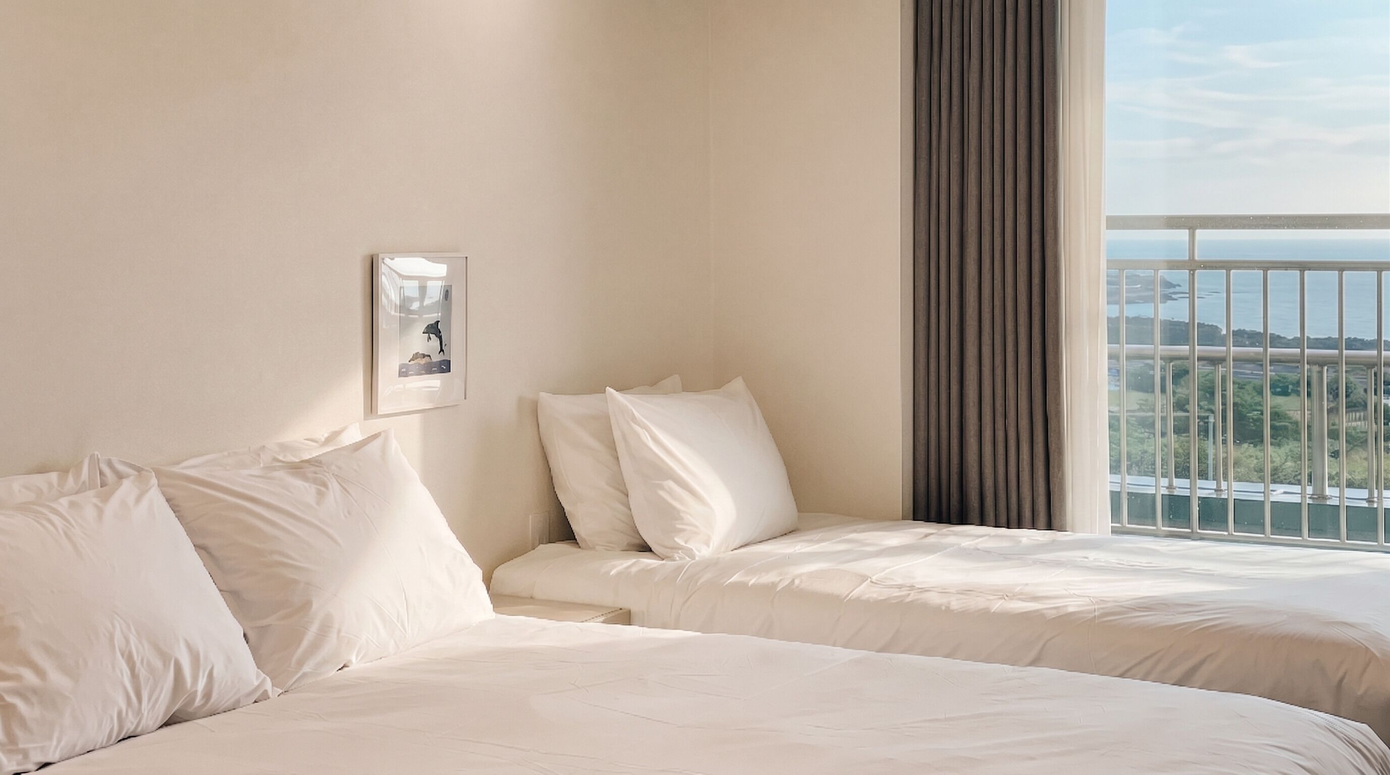 Quarto de hotel com duas camas, um quadro na parede e vista para o mar através de uma janela com cortinas.
