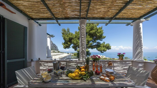 Mesa posta com frutas, doces e bebidas em terraço com vista para o mar.
