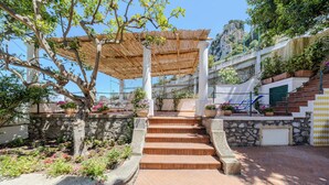 Uma escadaria de pedra leva a uma área coberta com pérgola, rodeada por vasos de plantas e com vista para uma paisagem montanhosa.