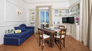 Uma sala de estar com um sofá azul, mesa de jantar de madeira, cadeiras, uma televisão e estantes com objetos decorativos.