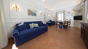 Uma sala de estar com um sofá azul, uma mesa de jantar com cadeiras, uma televisão e arandelas decorativas.