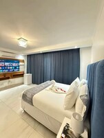 Quarto de hotel com cama, televisão fixada na parede, mesa de cabeceira com abajur e janela com cortinas.
