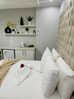 Cama bem arrumada com lençóis brancos e uma única rosa vermelha, mesa de cabeceira com uma chávena e bandeja, armário branco pequeno e prateleira na parede com objetos decorativos.