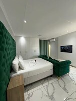 Quarto moderno com cabeceira verde estofada, cama branca, sofá verde, mesa de cabeceira de madeira e televisão fixada na parede.