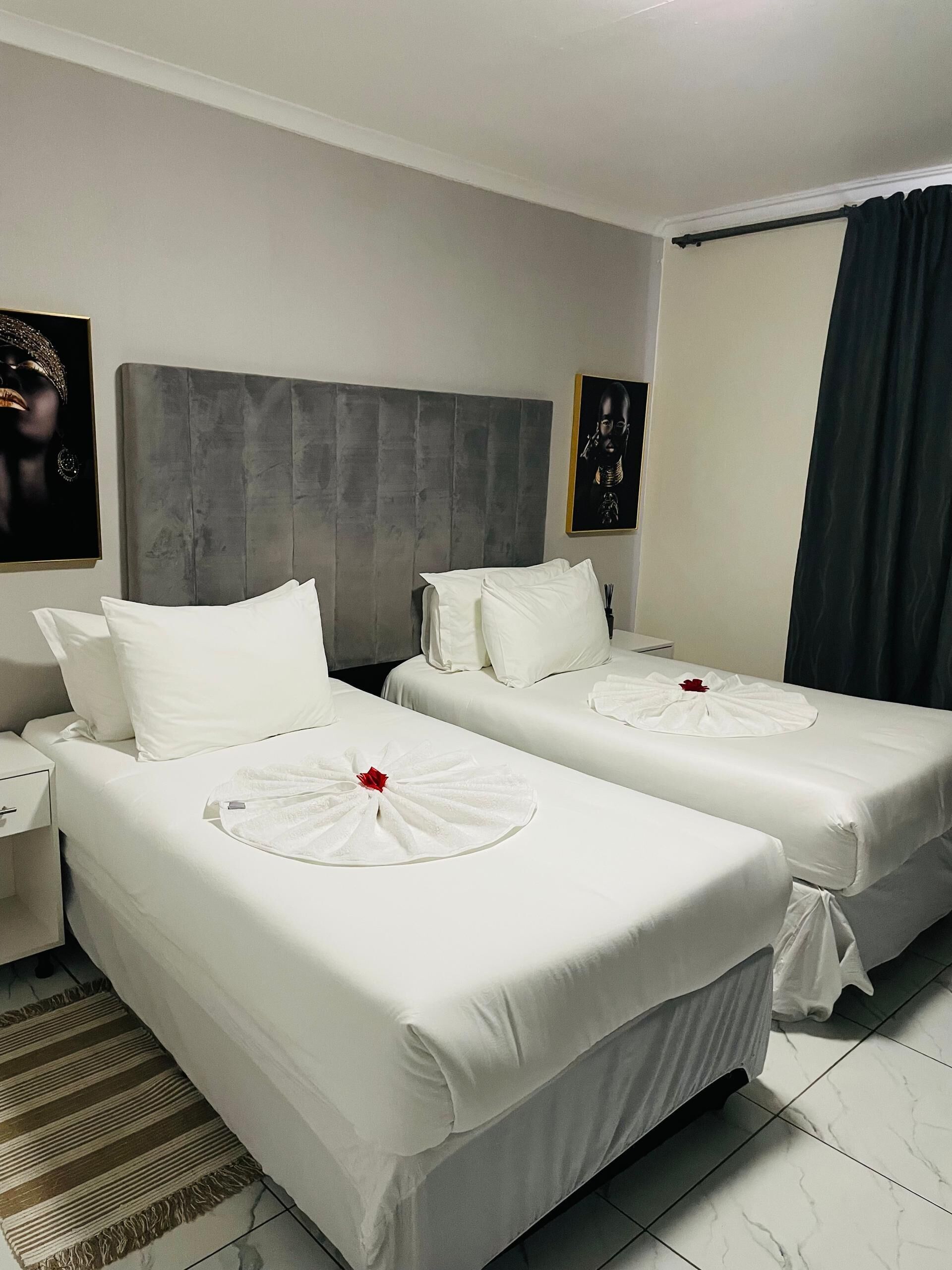 Quarto de hotel com duas camas, cabeceira cinza, roupa de cama branca, tapete listrado e cortina escura.
