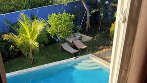 Vista de uma piscina através de uma janela, com uma cerca azul e vegetação ao fundo.