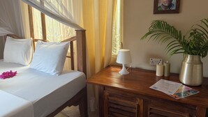Quarto de hotel com uma cama, mesa de cabeceira com abajur, um vaso com plantas e um quadro na parede.