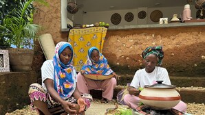 Três mulheres sentadas ao ar livre, preparando comida com uma panela de barro grande e diversos ingredientes.
