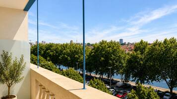 Uma varanda com planta em vaso, com vista para um estacionamento e um corpo d'água com árvores e edifícios ao fundo.