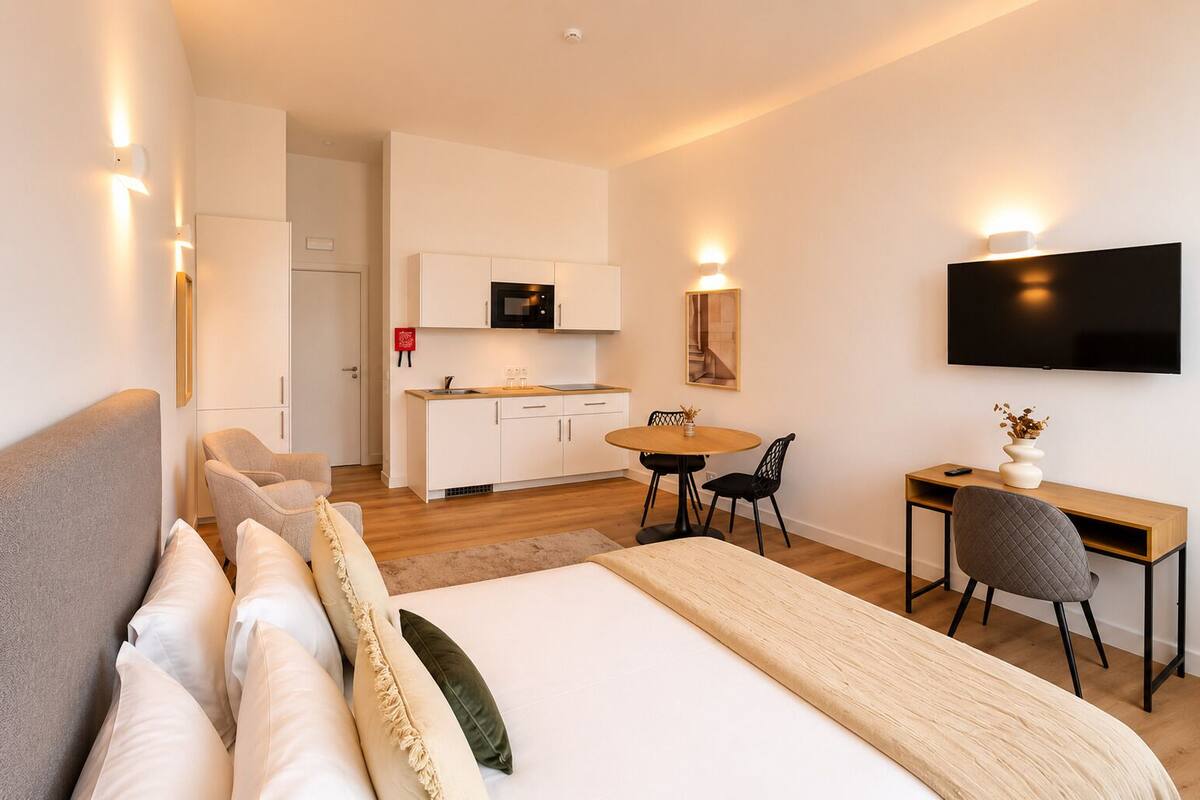 Quarto de hotel com cama, escrivaninha, cadeira, televisão e cozinha compacta.