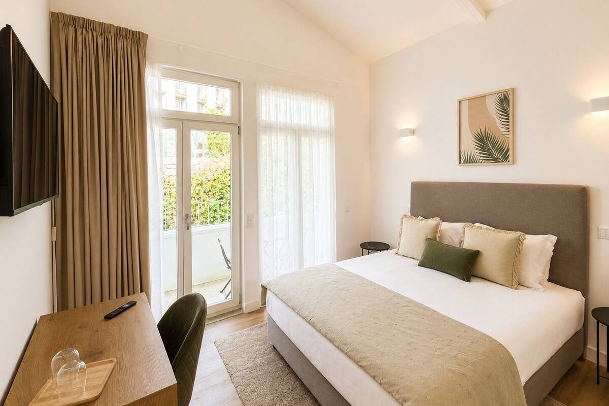 Quarto de hotel com cama, escrivaninha, cadeira, televisão e janela com cortinas.