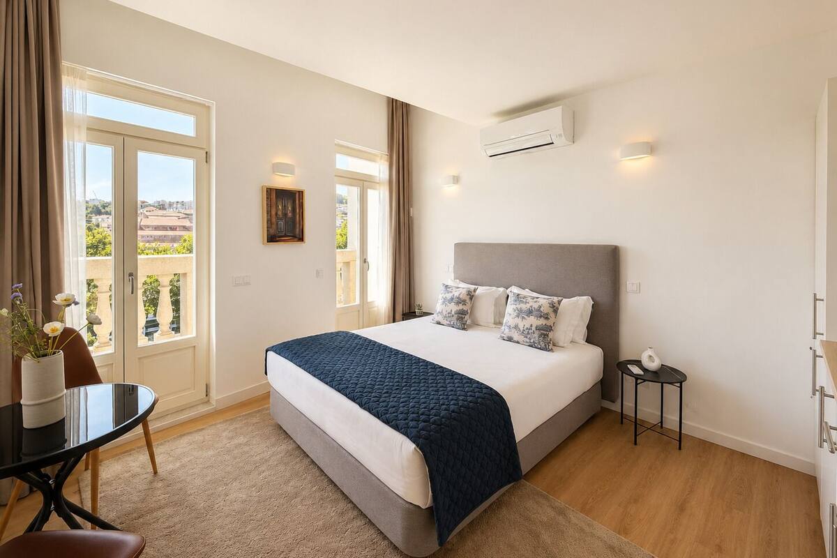 Quarto de hotel com cama grande, uma mesa pequena, uma cadeira e varanda com cortinas.