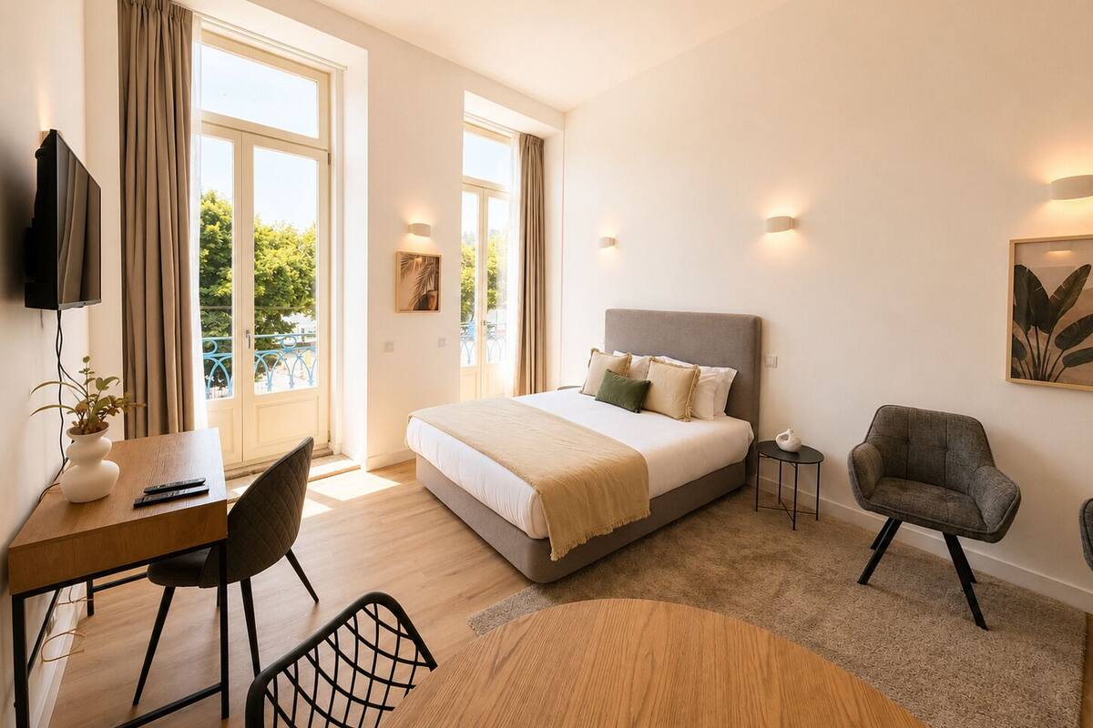 Quarto de hotel moderno com uma cama grande, uma escrivaninha, uma cadeira, uma mesinha e uma TV fixada na parede.