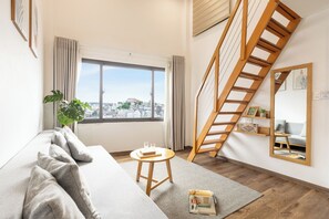 Uma sala de estar moderna com um sofá, uma pequena mesa redonda, uma planta, vista pela janela, uma escada de madeira e um espelho na parede.