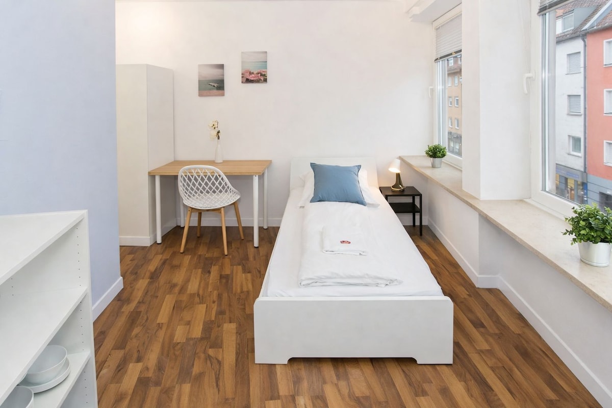 Quarto pequeno com uma cama de solteiro, escrivaninha, cadeira, mesa com abajur e janela com vista para edifícios.