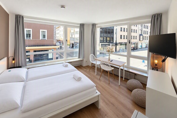 Quarto de hotel com cama grande, escrivaninha, duas cadeiras, televisão e vista para a cidade.