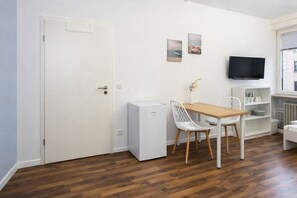 Quarto compacto com uma mesa de jantar de madeira, cadeiras brancas, um pequeno armário branco, uma televisão na parede e uma porta com puxador.