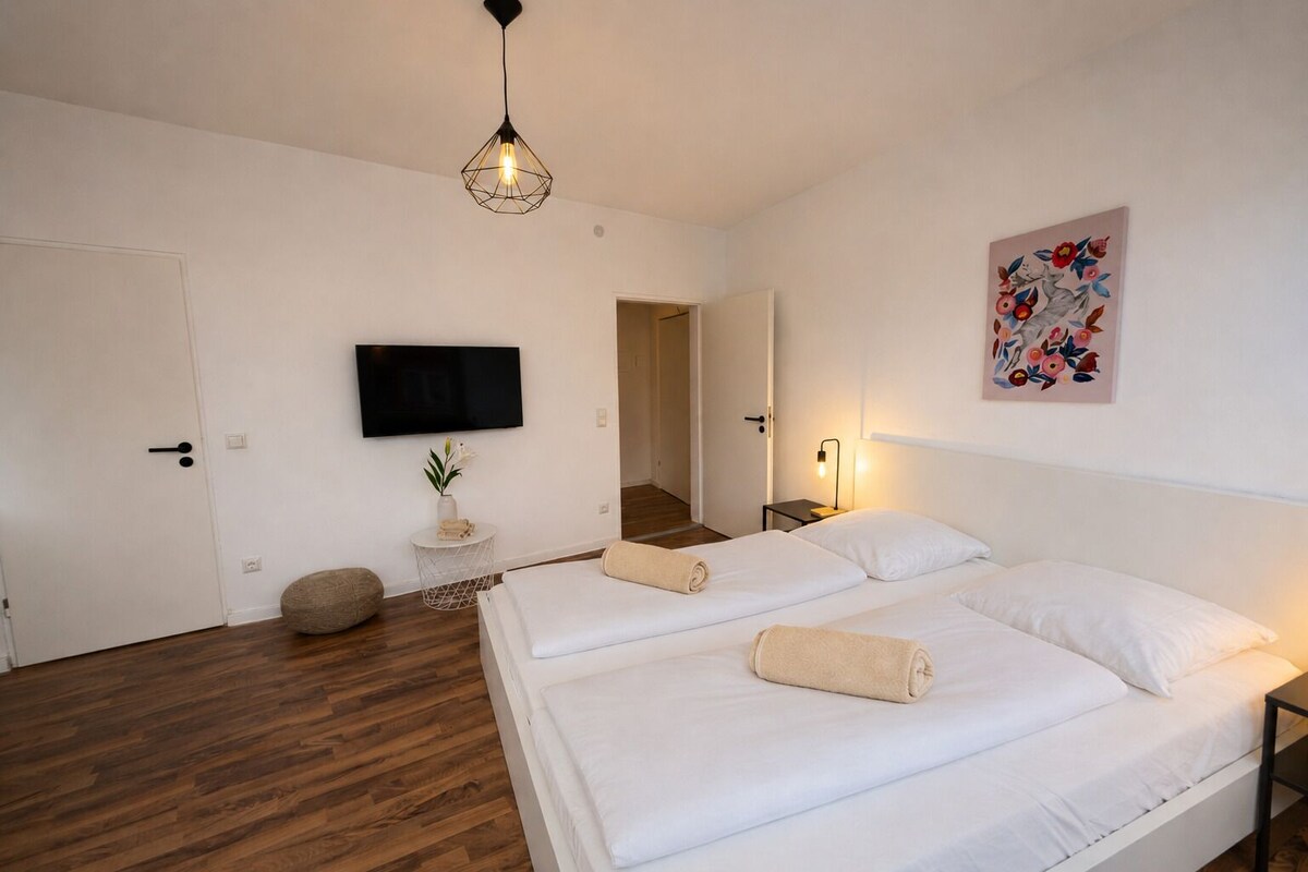 Quarto de hotel com duas camas, uma televisão, uma mesinha de apoio e um quadro na parede.