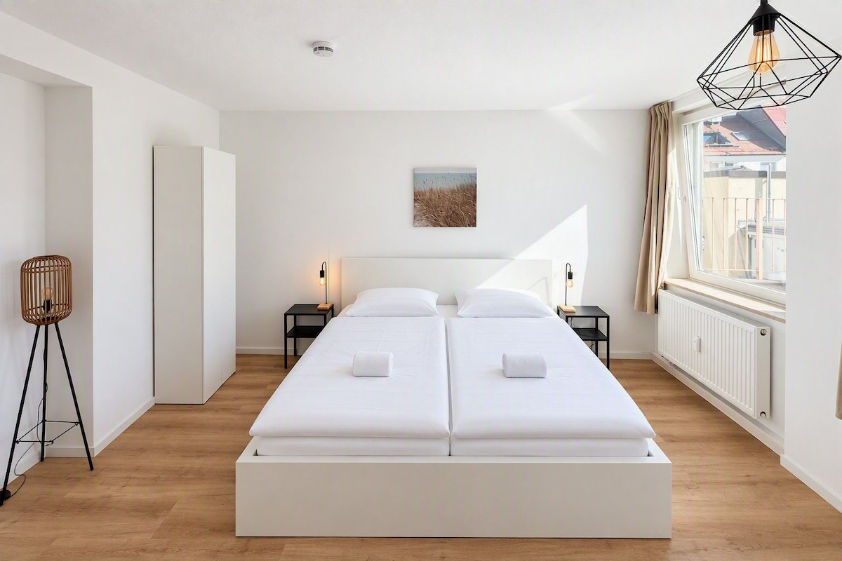 Quarto moderno com cama de casal, mesas de cabeceira, luminária de chão, radiador e janela com cortinas.