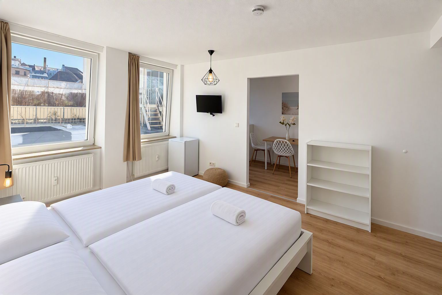 Quarto de hotel com duas camas, televisão, radiador, janela com vista e uma pequena mesa com cadeiras.