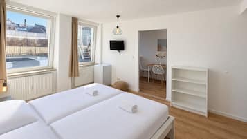 Quarto de hotel com duas camas, televisão, radiador, janela com vista e uma pequena mesa com cadeiras.