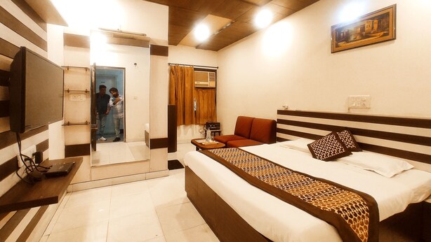 Quarto de hotel com cama, sofá, televisão, um quadro e banheiro visível através de uma porta aberta.