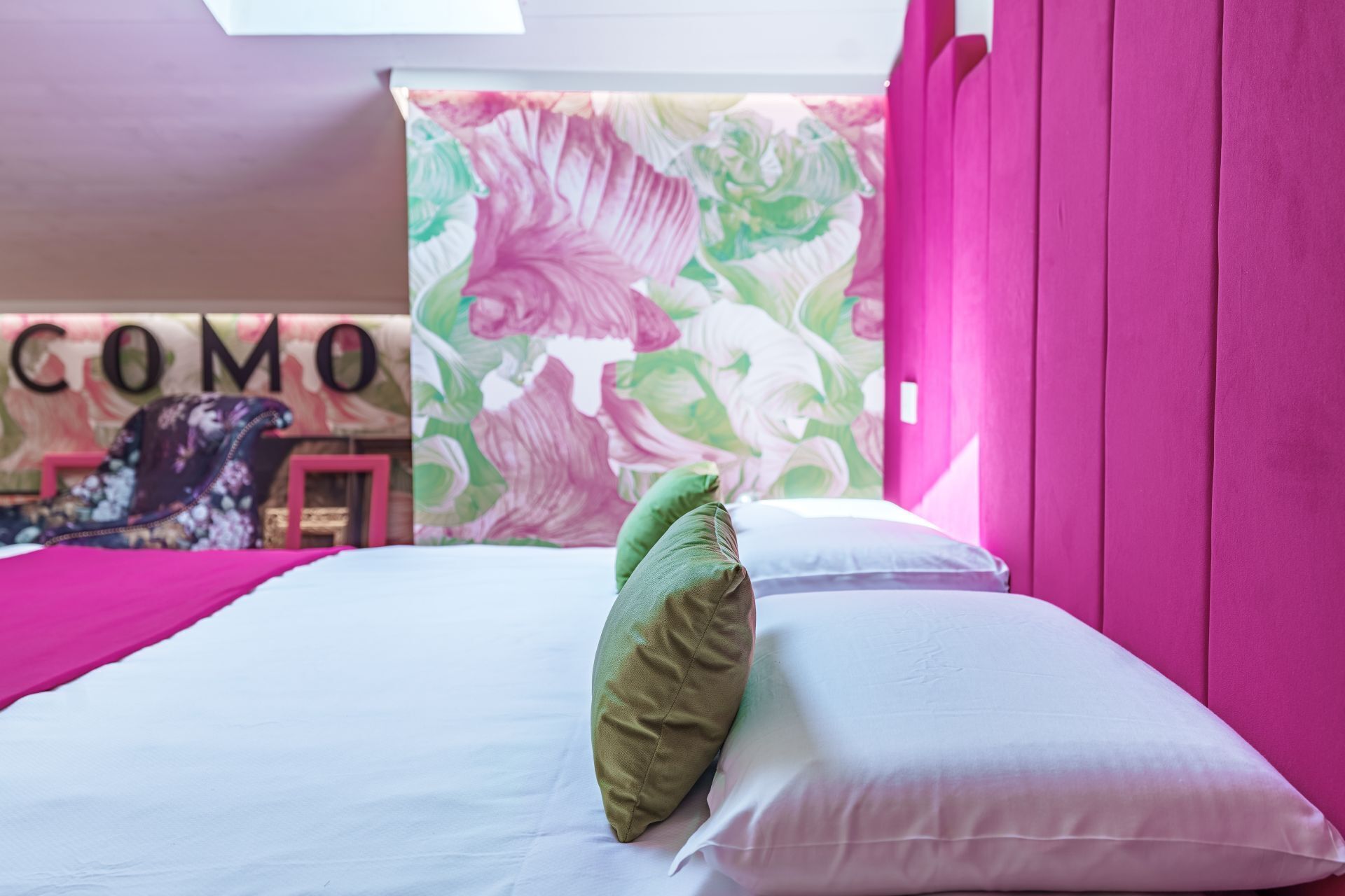 Cama com travesseiro verde e lençóis brancos, parede rosa e tapeçaria floral com a palavra 'COMO' parcialmente visível.