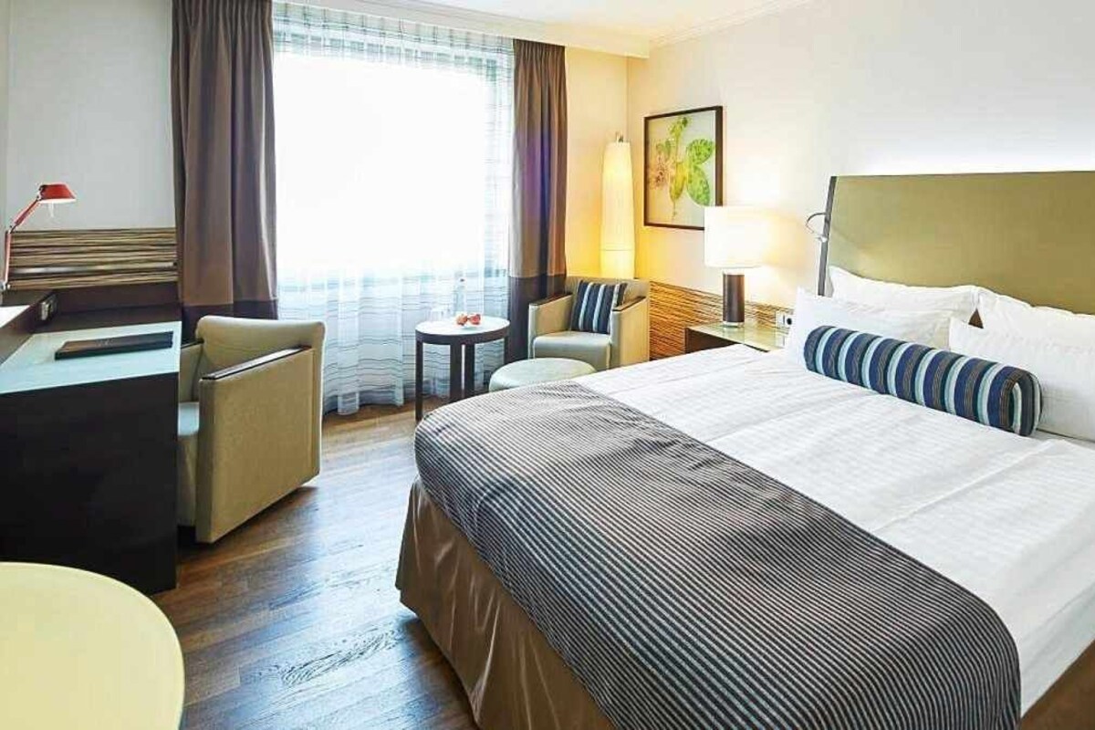 Quarto de hotel com uma cama grande, uma escrivaninha, uma cadeira, uma mesinha e uma janela com cortinas.
