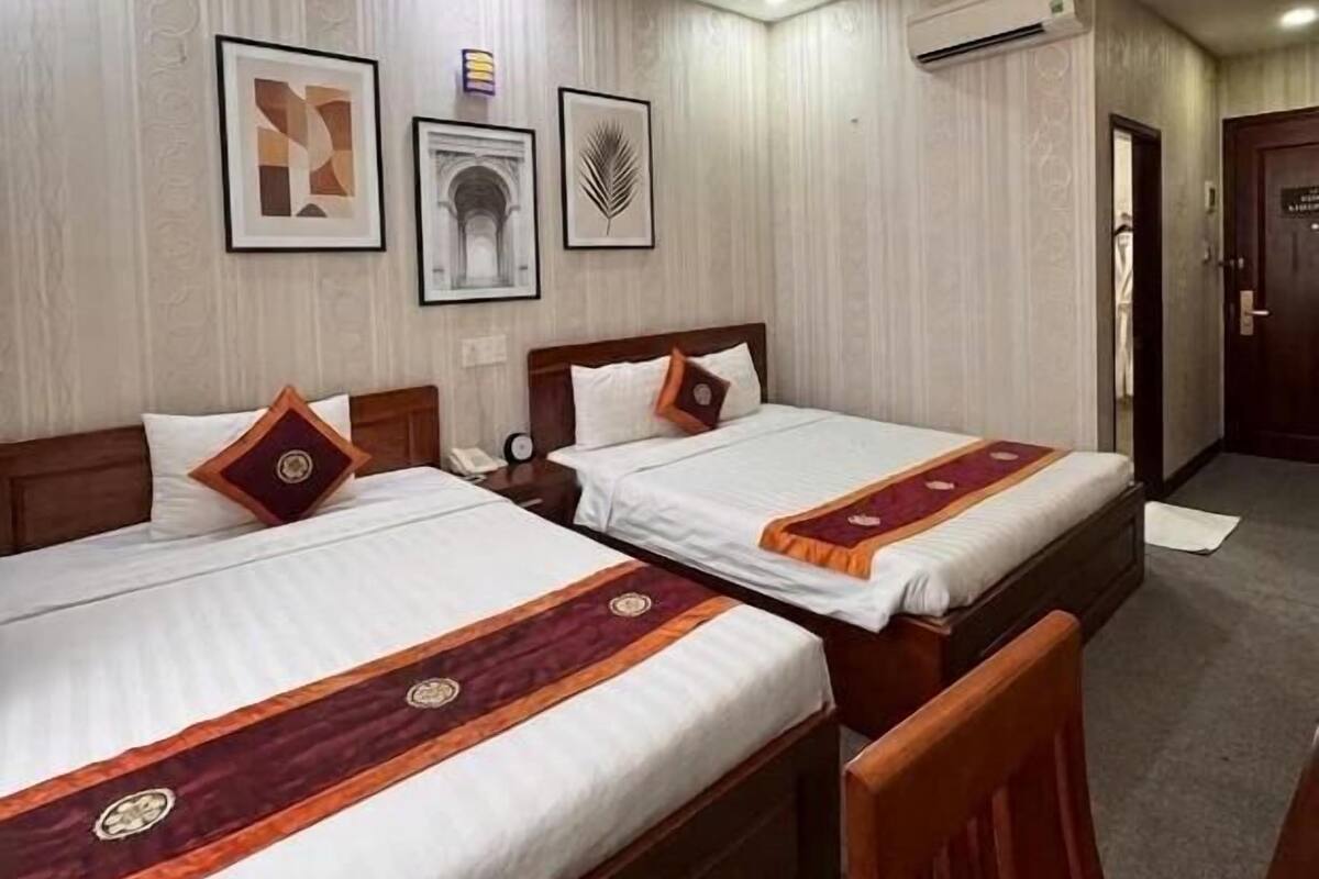 Quarto de hotel com duas camas, uma escrivaninha e uma cadeira. Há quadros nas paredes, um aparelho de ar condicionado e uma porta que dá para outro quarto.