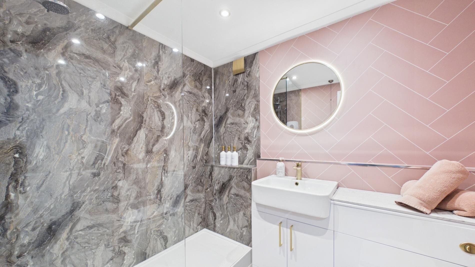 Casa de banho com parede de mármore, parede revestida a mosaico rosa, espelho redondo, lavatório branco e porta-toalhas.