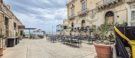 Área externa de café com mesas e cadeiras, vista para o mar e um edifício com uma placa.