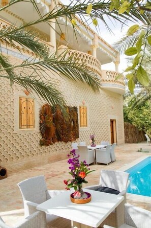 Área da piscina com mesa branca, cadeiras de vime e um vaso decorativo.
