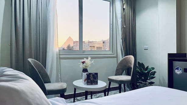 Quarto de hotel com uma cama, duas cadeiras, uma mesinha com um vaso e um livro, uma planta e vista para uma pirâmide pela janela.