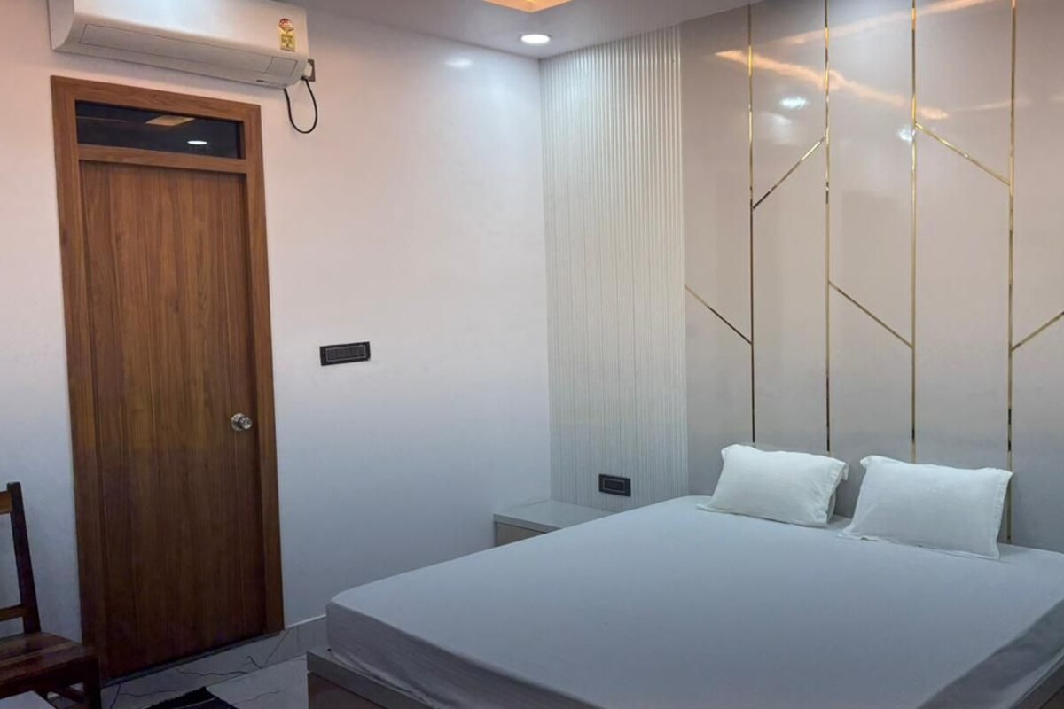 Quarto com porta de madeira, cama com roupa de cama branca e luminária fixada na parede.
