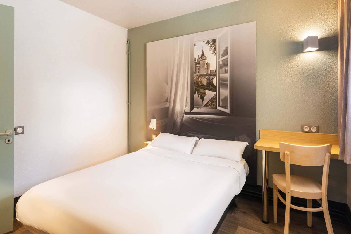 Quarto de hotel com uma cama, uma escrivaninha e uma cadeira. Há um quadro na parede acima da cama.