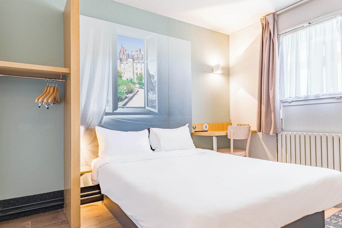 Quarto de hotel com cama, janela com cortinas, uma mesinha e um radiador.