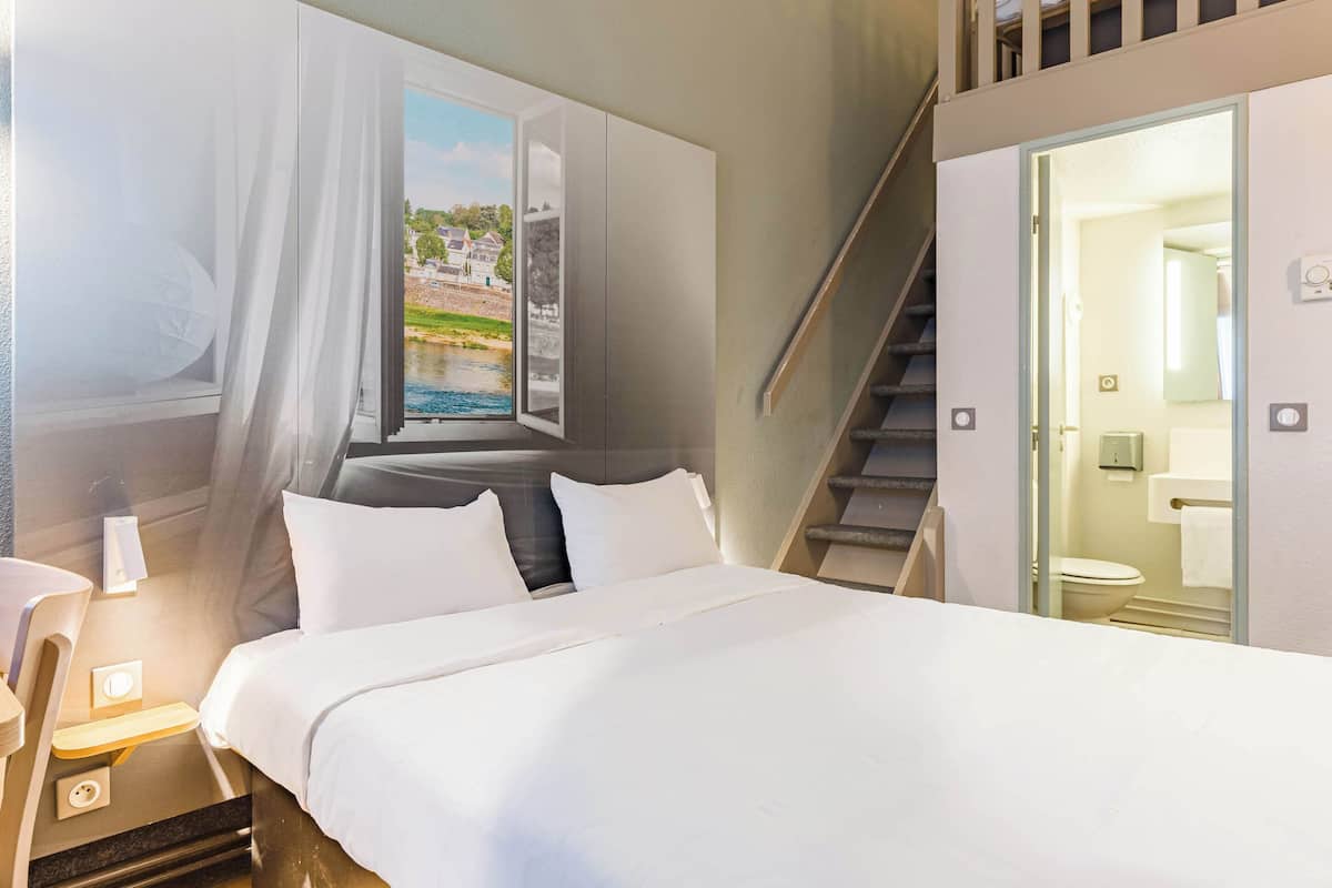 Quarto de hotel com uma cama, uma cadeira, uma janela com vista para um lago e edifícios, e uma escada que leva a um andar superior.