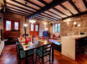 Interior rústico com vigas aparentes, paredes de pedra, área de jantar com mesa posta para quatro e uma cozinha compacta.