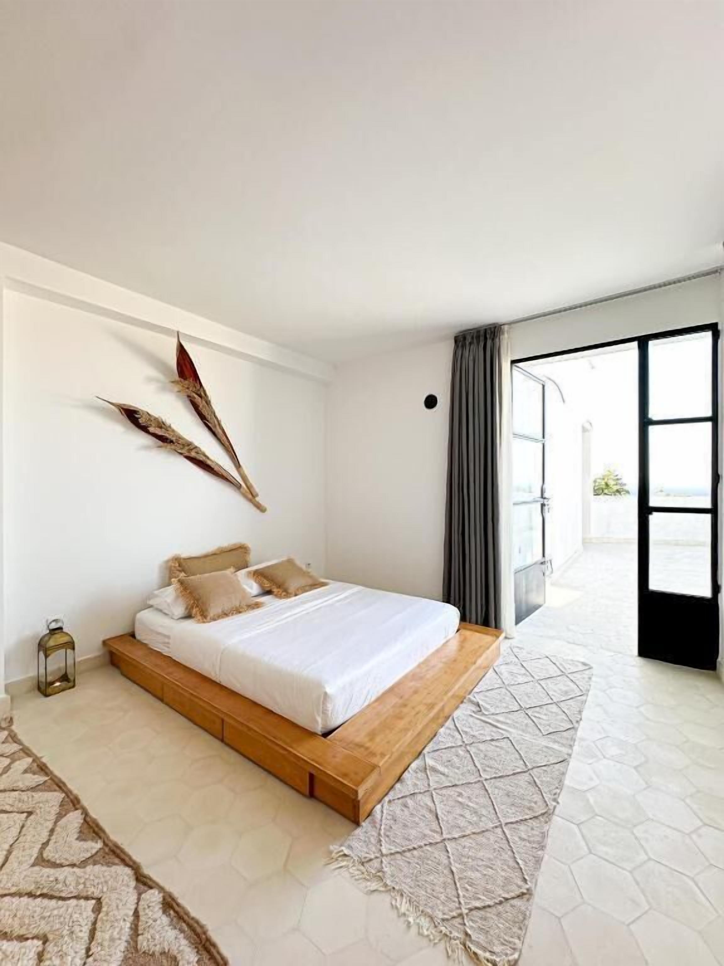 Quarto moderno com cama em plataforma de madeira, tapete com padrão geométrico, peça decorativa na parede e uma grande janela com vista.