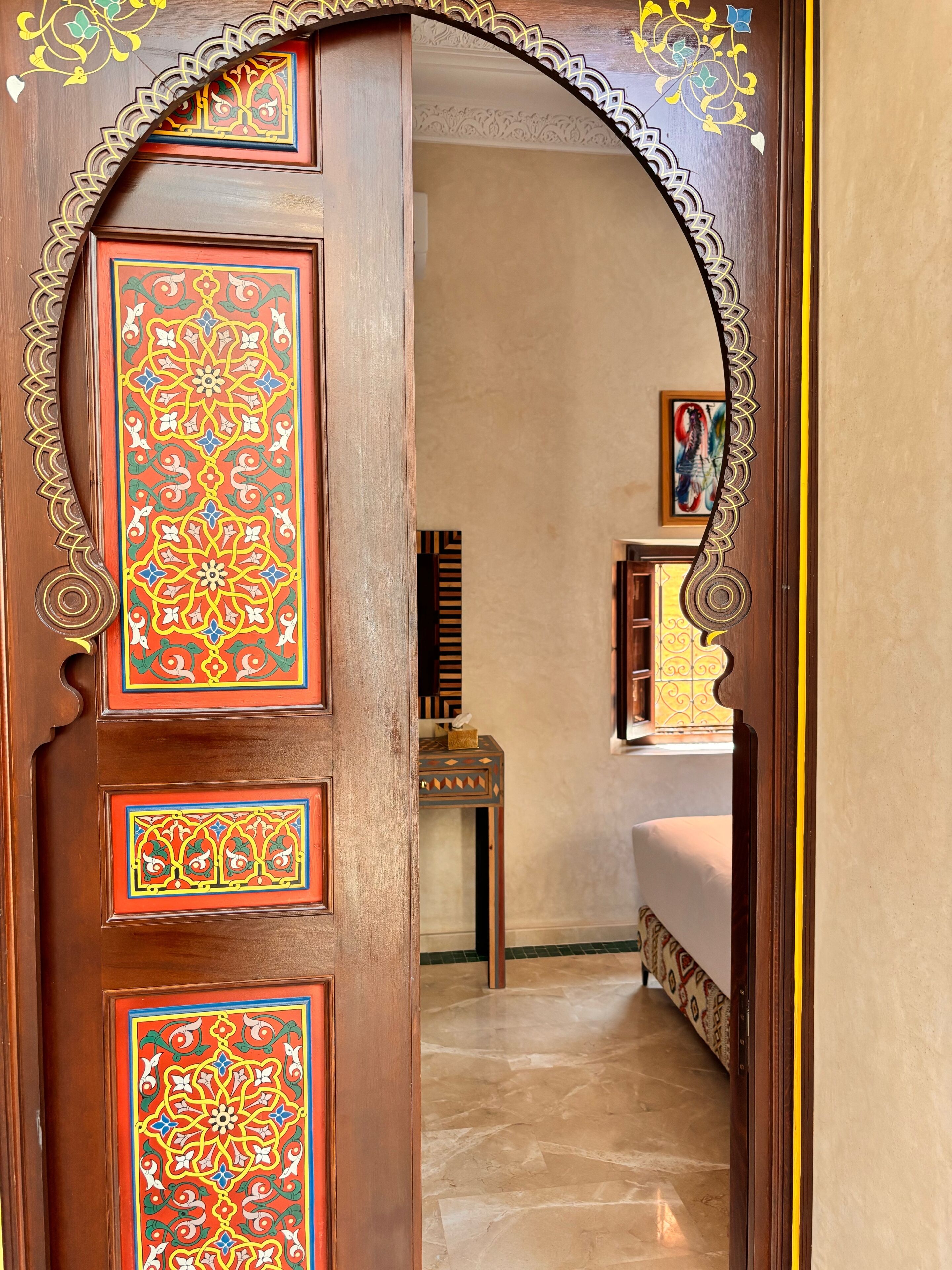Uma porta de madeira ornamentada com desenhos elaborados, dando acesso a um quarto com uma cama e uma pequena mesa.