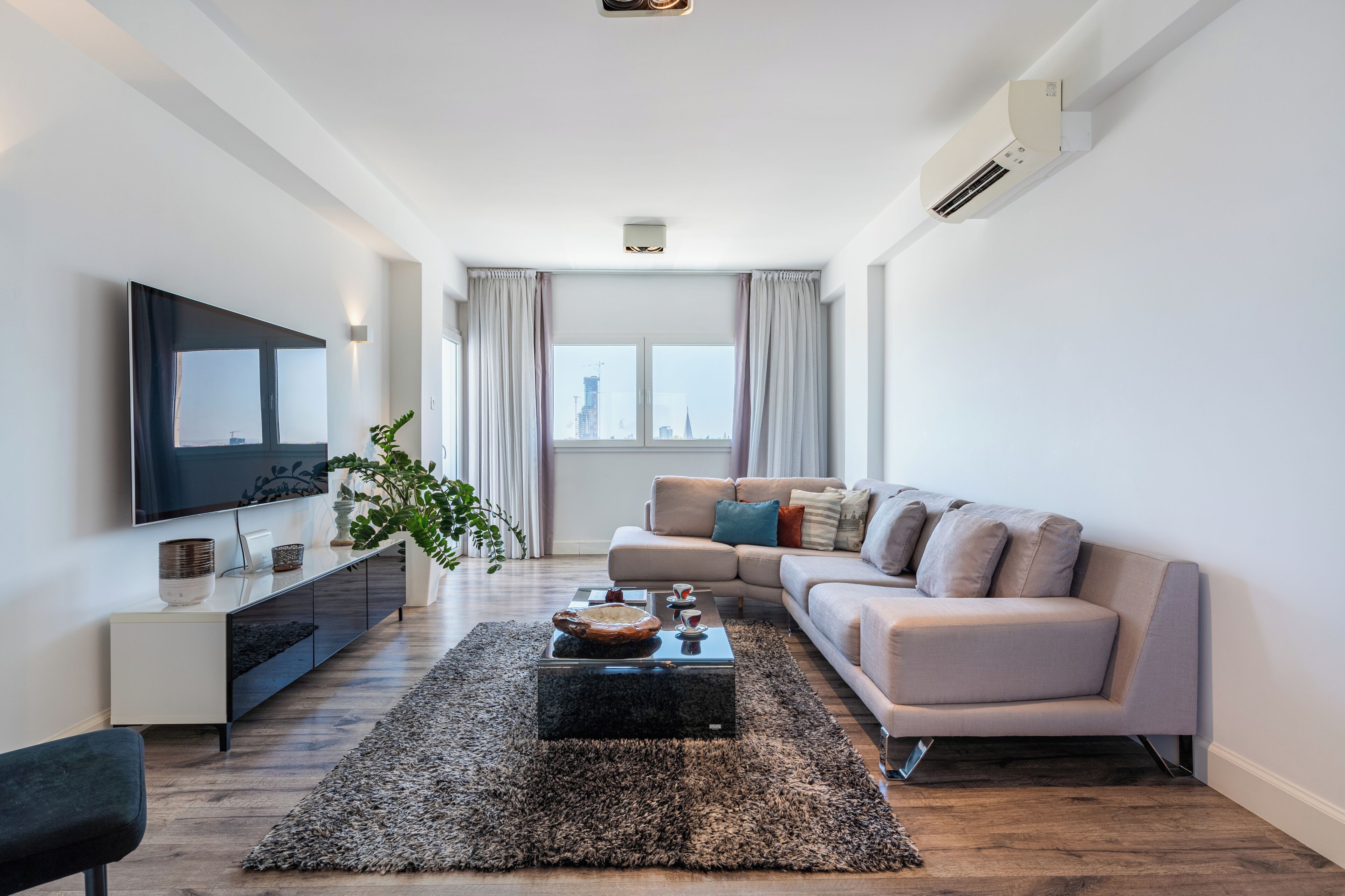 Uma sala de estar moderna com televisão de tela plana, sofá de canto, mesa de centro e uma janela grande com cortinas.