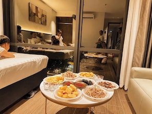 Quarto de hotel com uma cama, uma mesa de jantar e vários alimentos, e um espelho refletindo o ambiente.