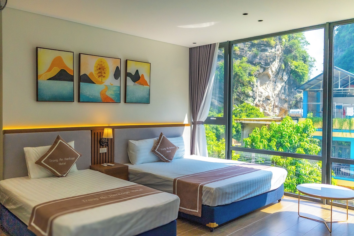 Quarto de hotel com duas camas, uma mesa pequena e uma janela grande que oferece vista para áreas verdes e um edifício.