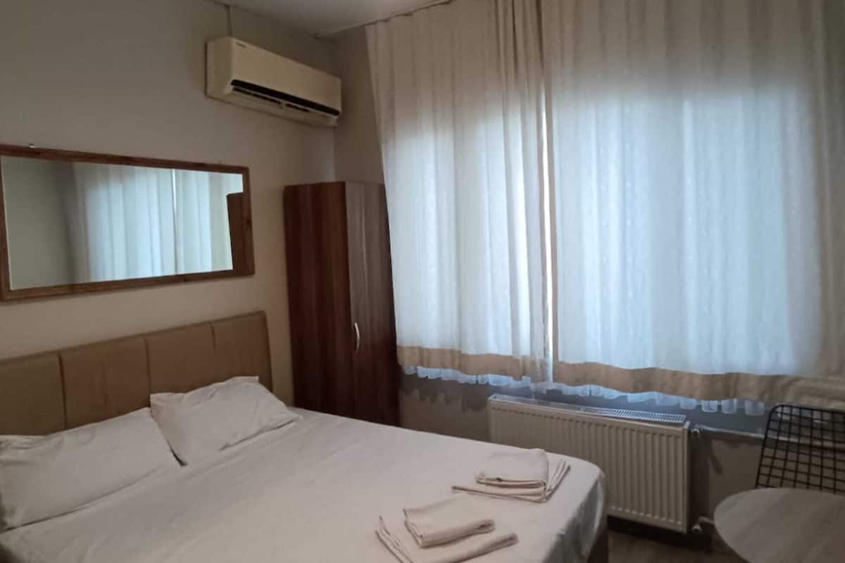 Quarto de hotel com cama, espelho, guarda-roupa, radiador, televisão e janela com cortinas.