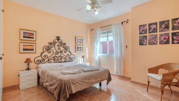 Quarto com cama, mesa de cabeceira, cadeira, ventilador de teto e obras de arte nas paredes.