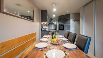 Uma sala de jantar moderna com uma mesa de madeira posta para seis pessoas, rodeada por cadeiras e um espelho grande na parede.