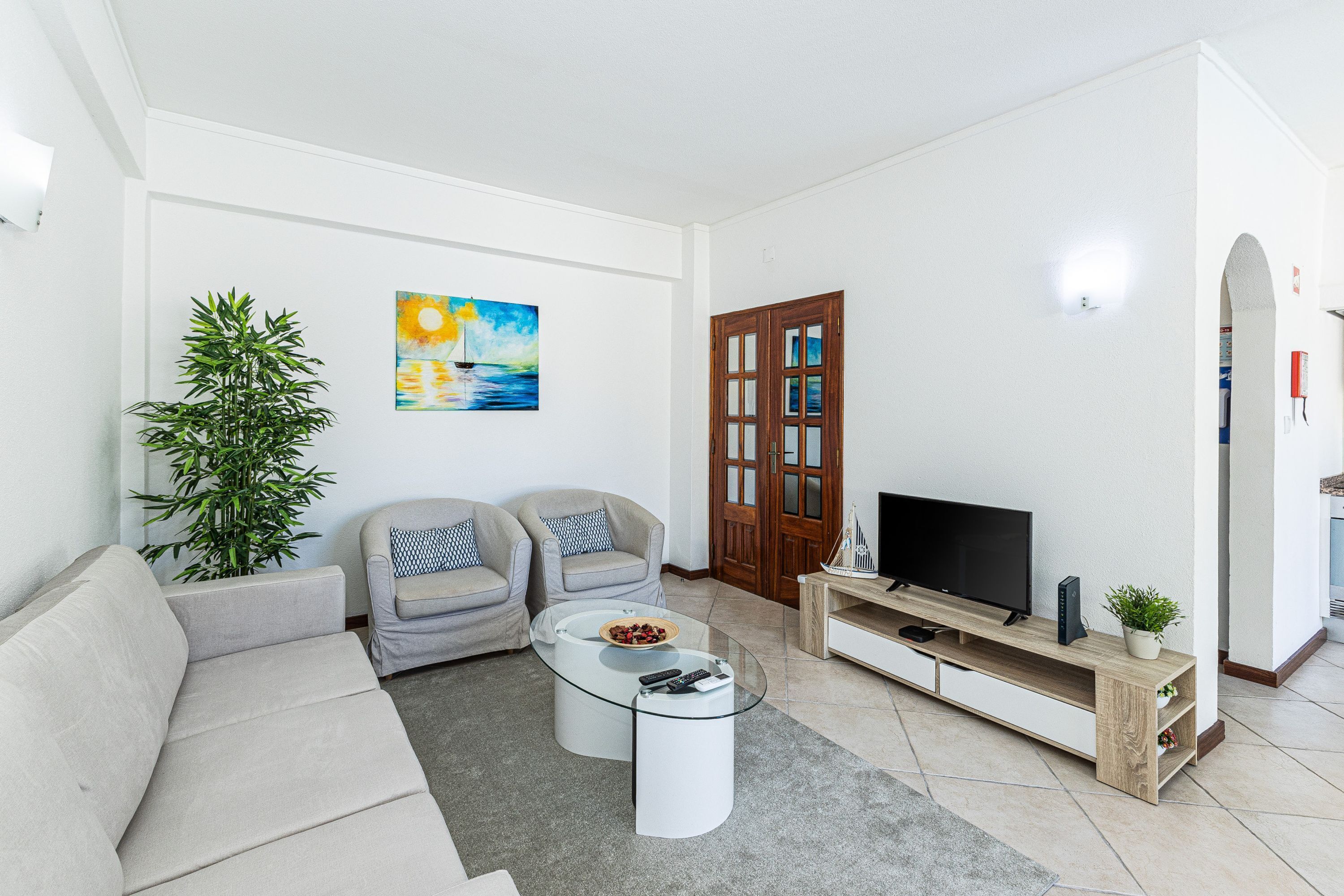 Uma sala de estar com sofá, duas poltronas, mesa de centro, televisão sobre um suporte de madeira, quadro na parede e planta em vaso.