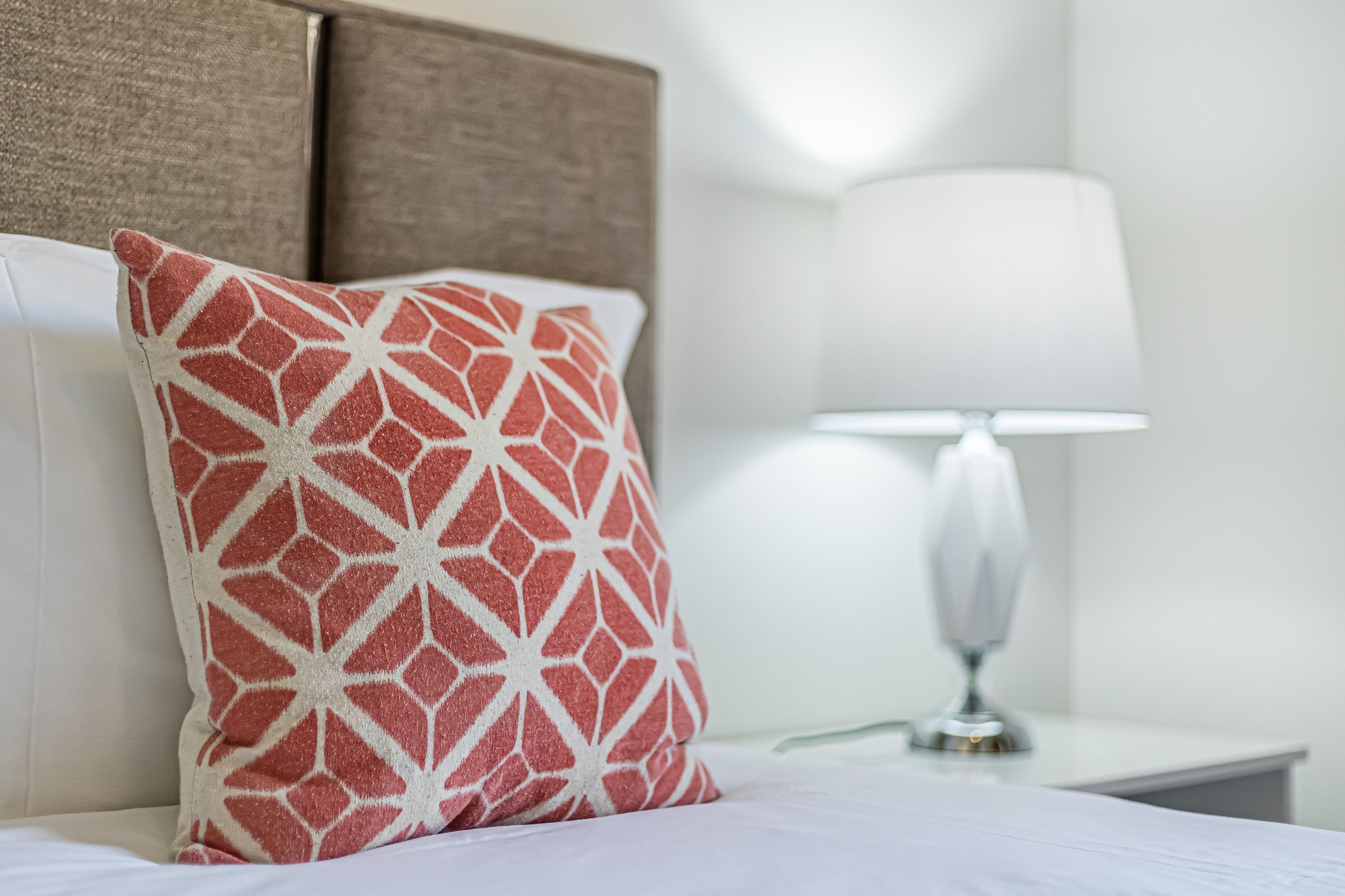 Cama bem arrumada com travesseiro estampado, luminária de cabeceira branca e cabeceira.