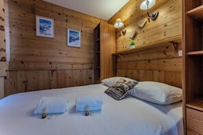 Quarto com painéis de madeira, uma cama, duas toalhas, um travesseiro, uma luminária fixada na parede e duas imagens emolduradas.